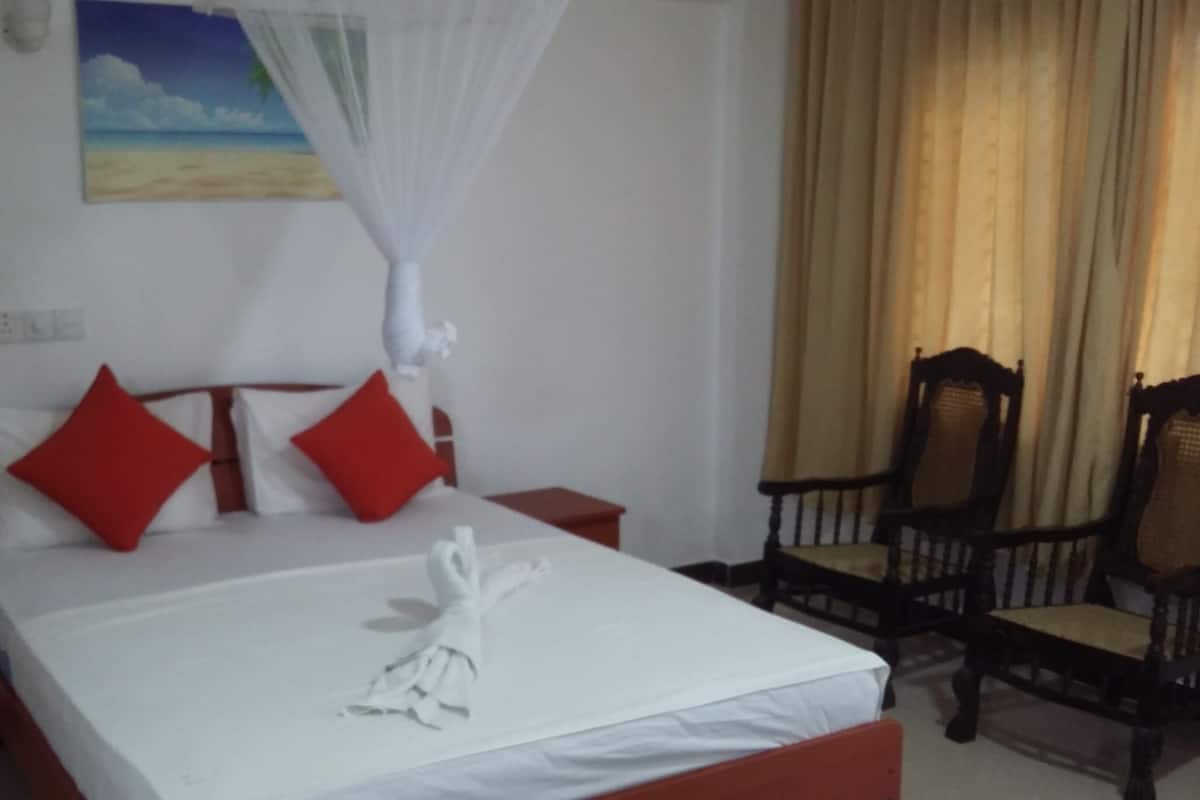 Quarto de hotel com uma cama, duas almofadas vermelhas, um edredom branco, uma toalha branca, um quadro de uma praia e uma cadeira.