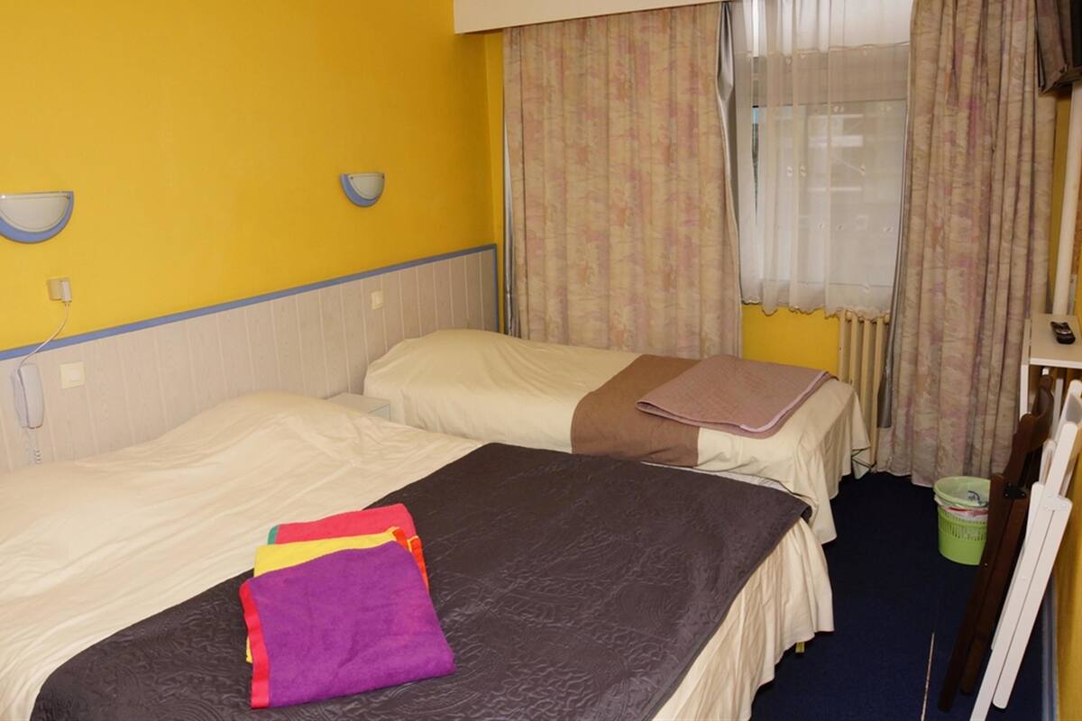 Quarto de hotel com duas camas, uma mesa pequena, uma cadeira, janela com cortinas e radiador.