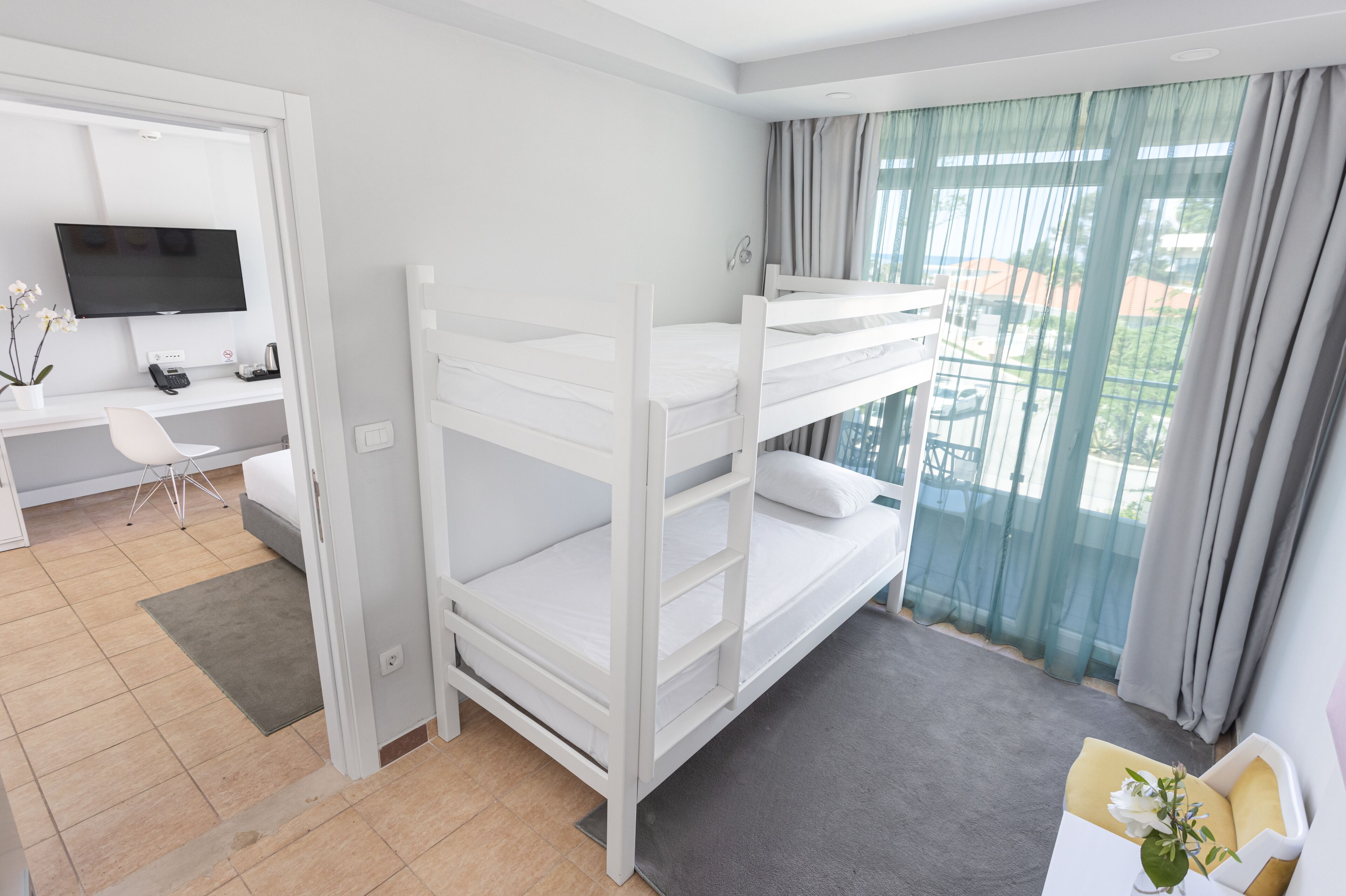 Quarto de hotel moderno com beliche, escrivaninha com cadeira, televisão e uma janela grande com cortinas.