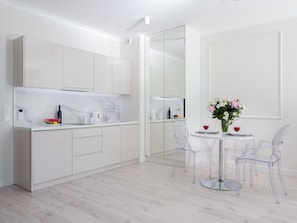 Cozinha moderna com armários brancos, bancada de mármore e área de refeições com cadeiras transparentes e um vaso de flores.