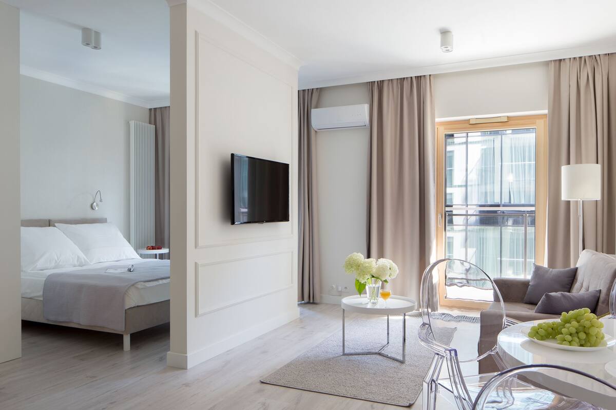 Quarto de hotel moderno com cama, televisão, área de refeições com mesa e cadeiras, e uma grande janela com cortinas.
