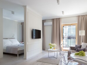 Quarto de hotel moderno com cama, televisão, área de refeições com mesa e cadeiras, e uma grande janela com cortinas.