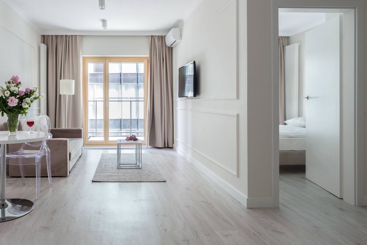 Quarto de hotel moderno com uma janela grande, televisão de tela plana fixada na parede, área de jantar com mesa e cadeiras, e uma cama no quarto adjacente.