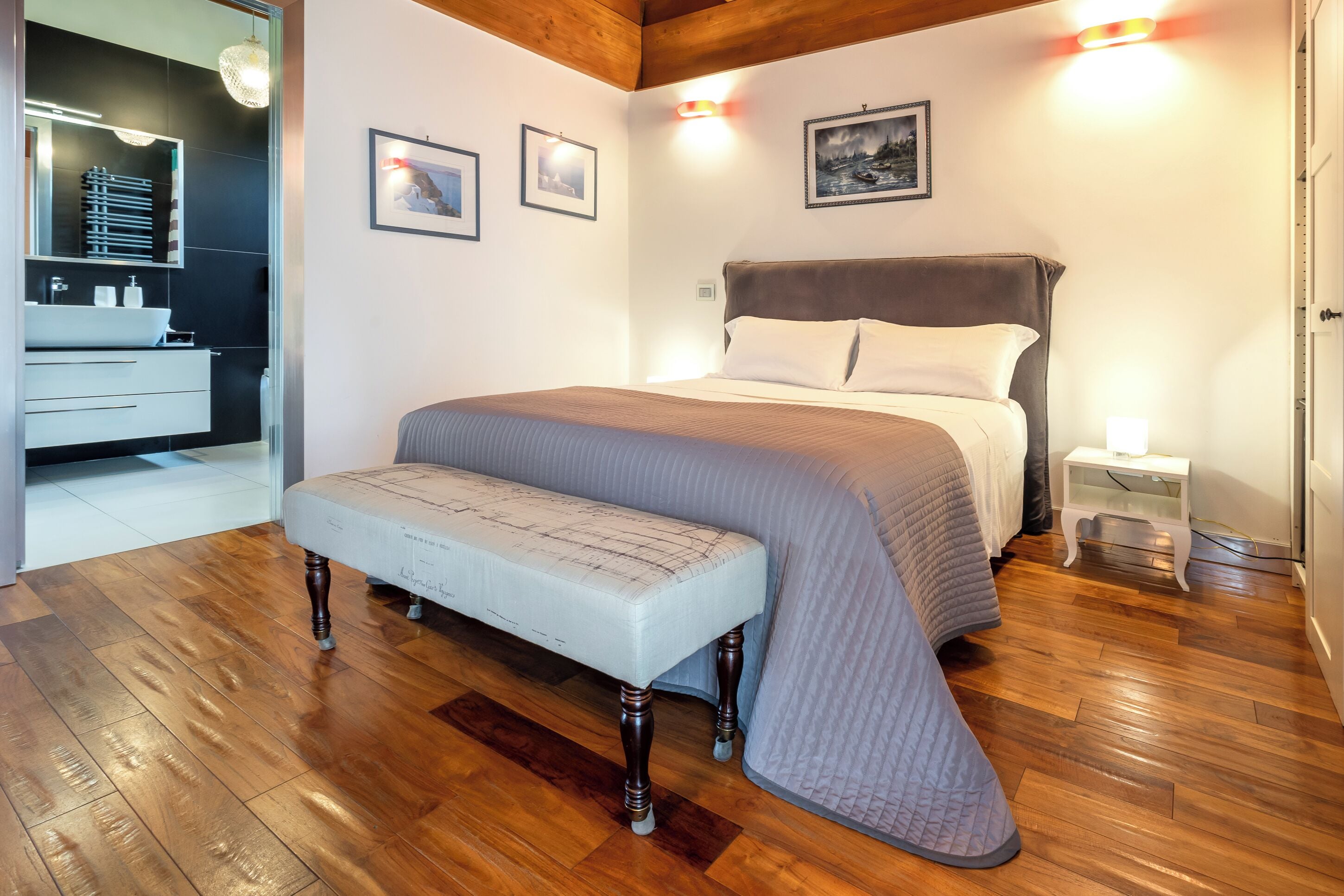 Quarto com cama, banqueta, mesa de cabeceira e uma pequena mesa de apoio.