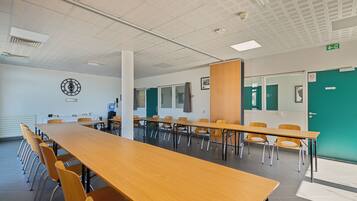 Uma sala de conferências com uma longa mesa de madeira e várias cadeiras, teto branco com padrão quadriculado, e uma porta verde ao fundo.