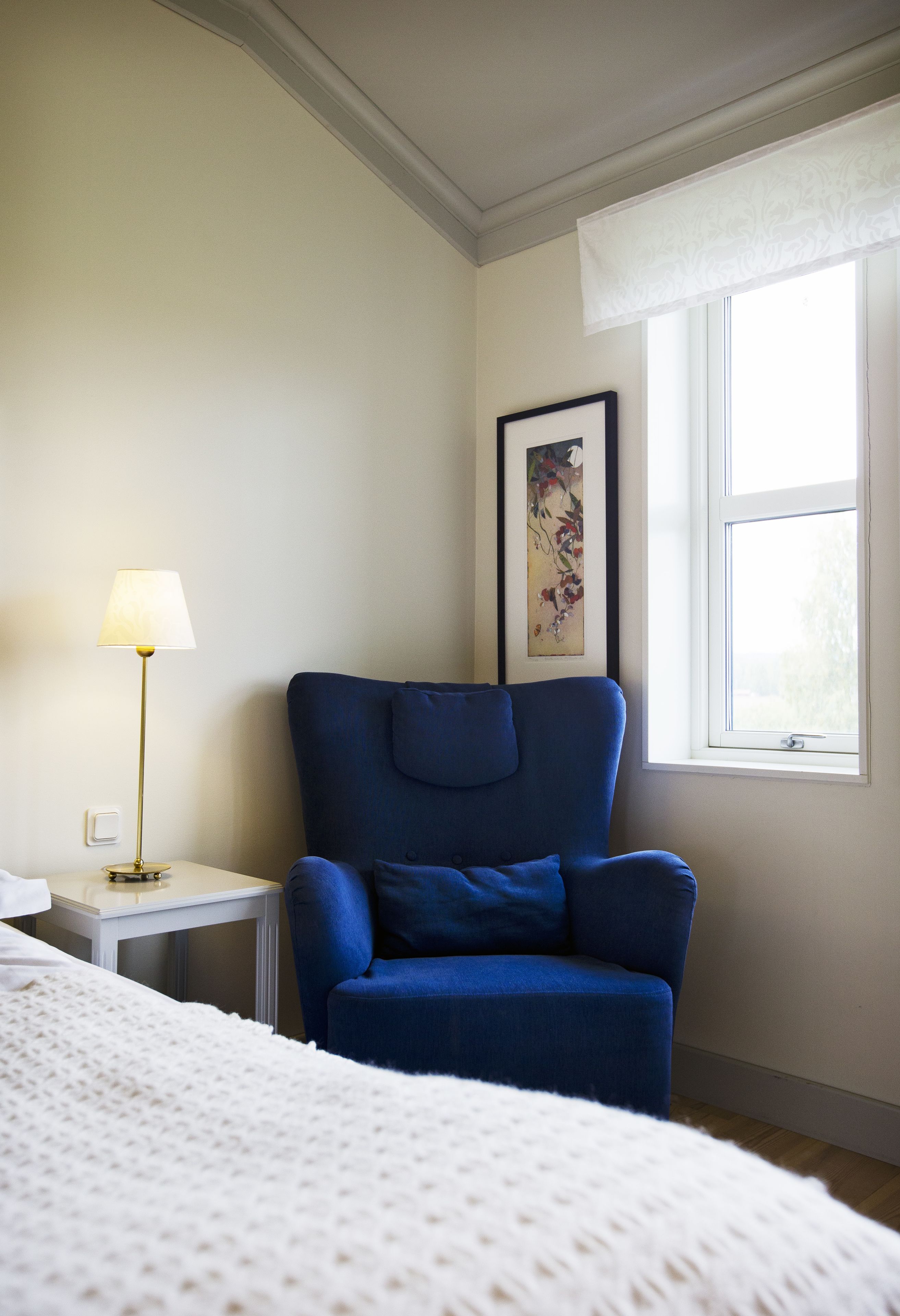 Quarto com poltrona azul, mesa de cabeceira branca com abajur e uma obra de arte emoldurada na parede.