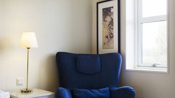 Quarto com poltrona azul, mesa de cabeceira branca com abajur e uma obra de arte emoldurada na parede.