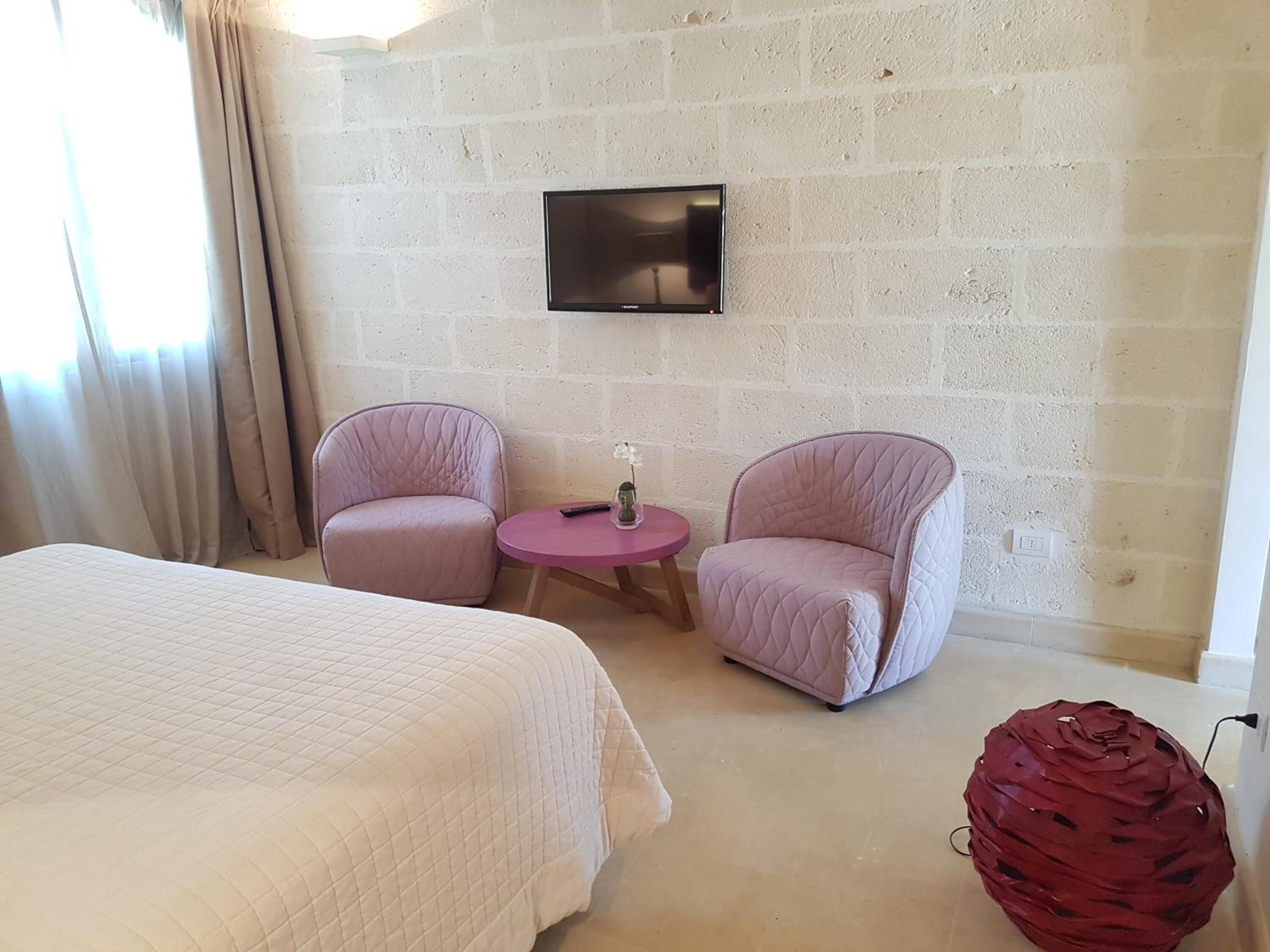 Quarto de hotel com cama, duas poltronas, uma mesa pequena, televisão presa a uma parede de tijolos e um objeto decorativo vermelho.