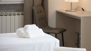Cama bem arrumada com toalhas brancas dobradas, uma cadeira estofada cinza, uma pequena escrivaninha com luminária e um radiador ao fundo.