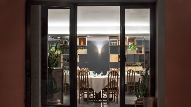 Uma área de refeições aberta com uma mesa posta para quatro pessoas, rodeada de cadeiras, e prateleiras com diversos objetos ao fundo.