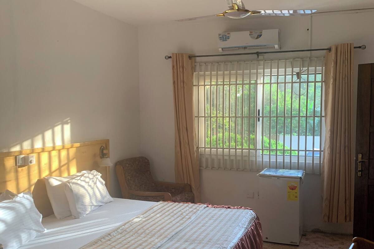 Quarto de hotel com cama, uma cadeira, janela com cortinas, ar condicionado e porta.