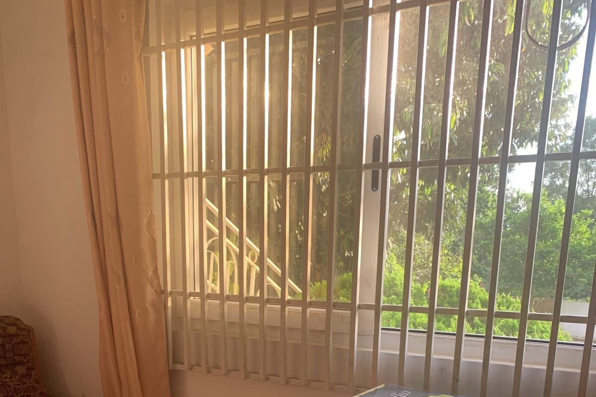 Quarto com sofá estampado, cama com manta listrada, frigorífico branco pequeno e janela com grades de metal.