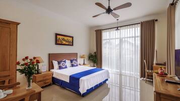 Quarto de hotel com cama, escrivaninha, cadeira, mesa de cabeceira, luminária, planta, quadro e ventilador de teto.