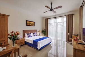 Quarto de hotel com cama, escrivaninha, cadeira, mesa de cabeceira, luminária, planta, quadro e ventilador de teto.