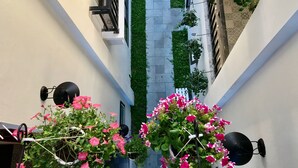 Uma viela estreita com paredes brancas, caminho de azulejos e plantas pendentes com flores cor-de-rosa.