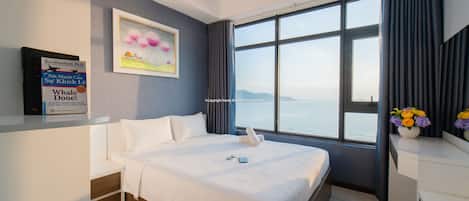 Quarto de hotel com uma cama grande, mesa de cabeceira, escrivaninha, janela com vista para o mar e um quadro na parede.