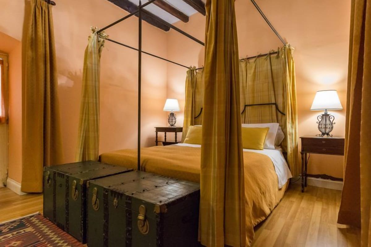 Uma cama com dossel e cortina, roupa de cama amarela, duas mesas de cabeceira com abajures e um baú antigo no canto.