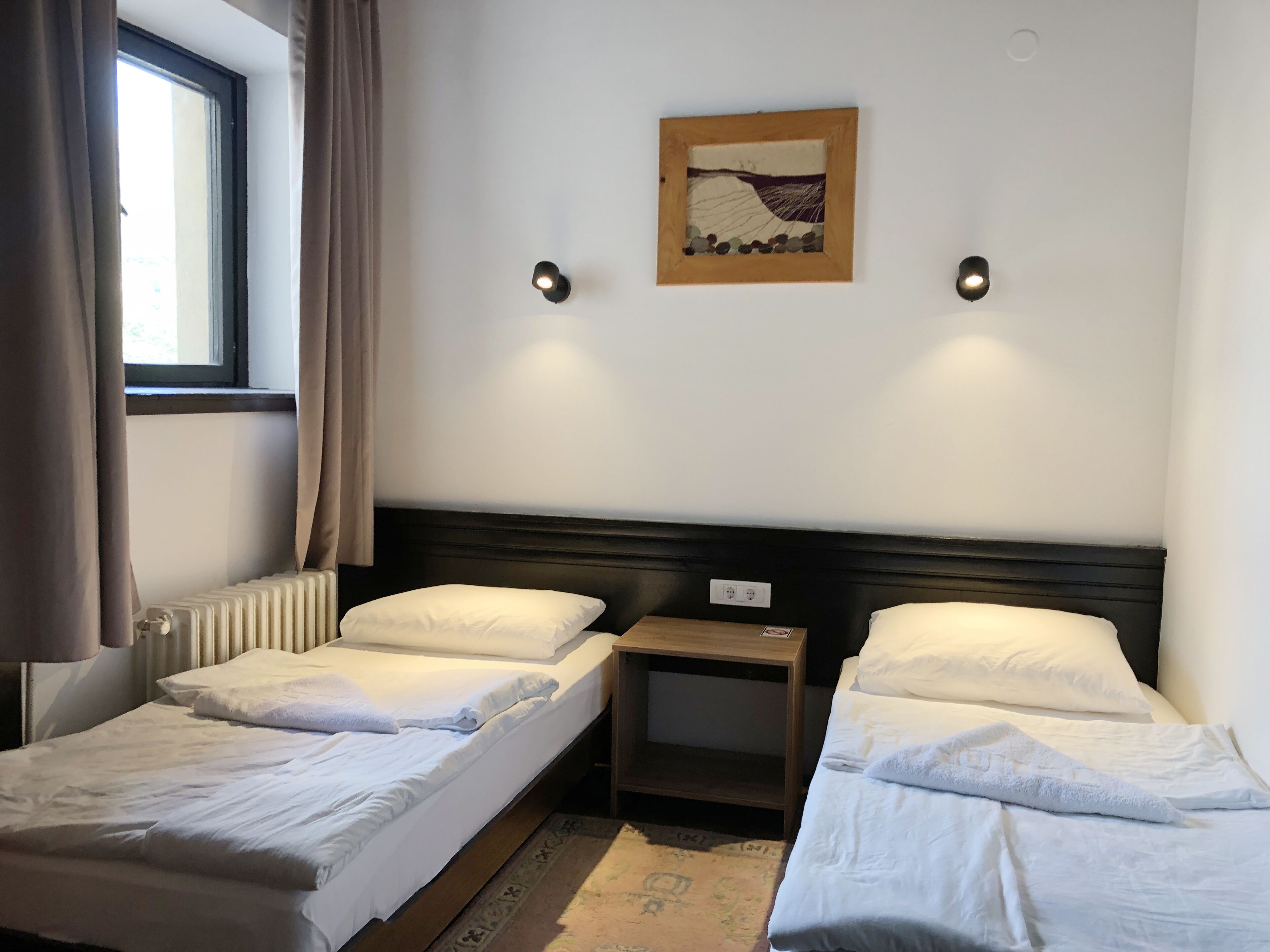 Quarto de hotel com duas camas de solteiro, uma pequena mesa de cabeceira, um radiador, janela com cortinas e um quadro na parede.