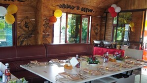 Restaurante com uma longa mesa de jantar posta para uma refeição, rodeado por paredes de madeira e grandes janelas.