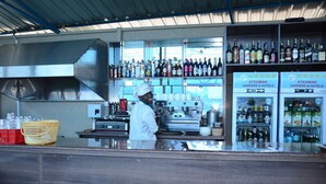 Uma área de bar com um chefe preparando bebidas, prateleiras com diversos frascos e uma geladeira com a etiqueta 'STEDMAK GARDENS & HOTELS'.