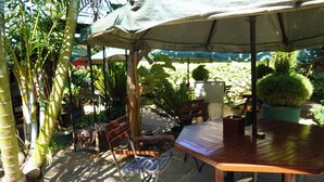 Um pátio externo com mesa e cadeiras de madeira, rodeado por plantas em vasos e um grande guarda-sol que oferece sombra.