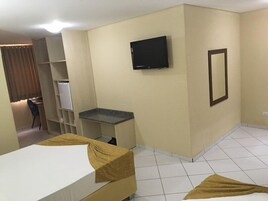 Quarto de hotel com cama, escrivaninha, cadeira, televisão e espelho.