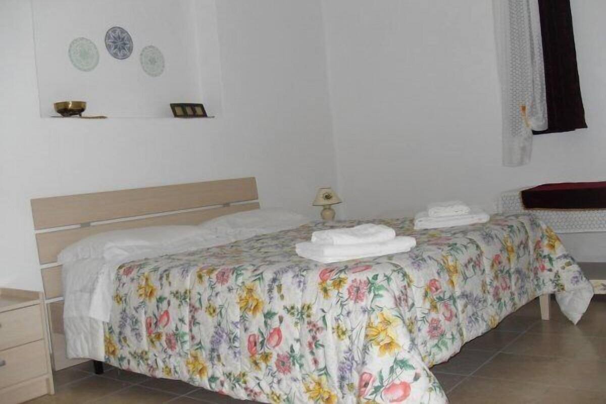 Quarto com cama, mesa de cabeceira, abajur e janela com cortinas.