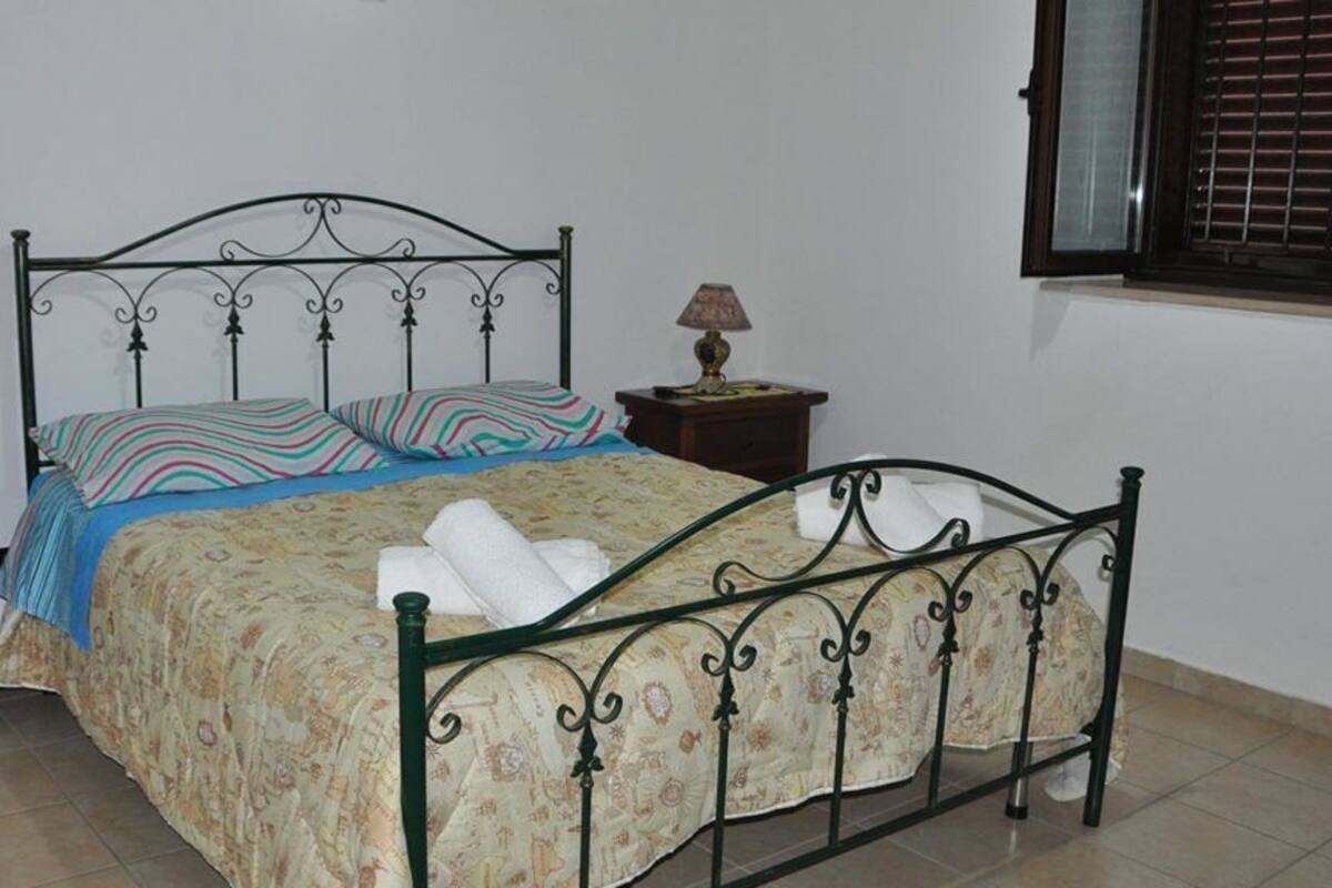Uma cama de solteiro com estrutura de metal, um criado-mudo de madeira com abajur e uma janela com persianas.