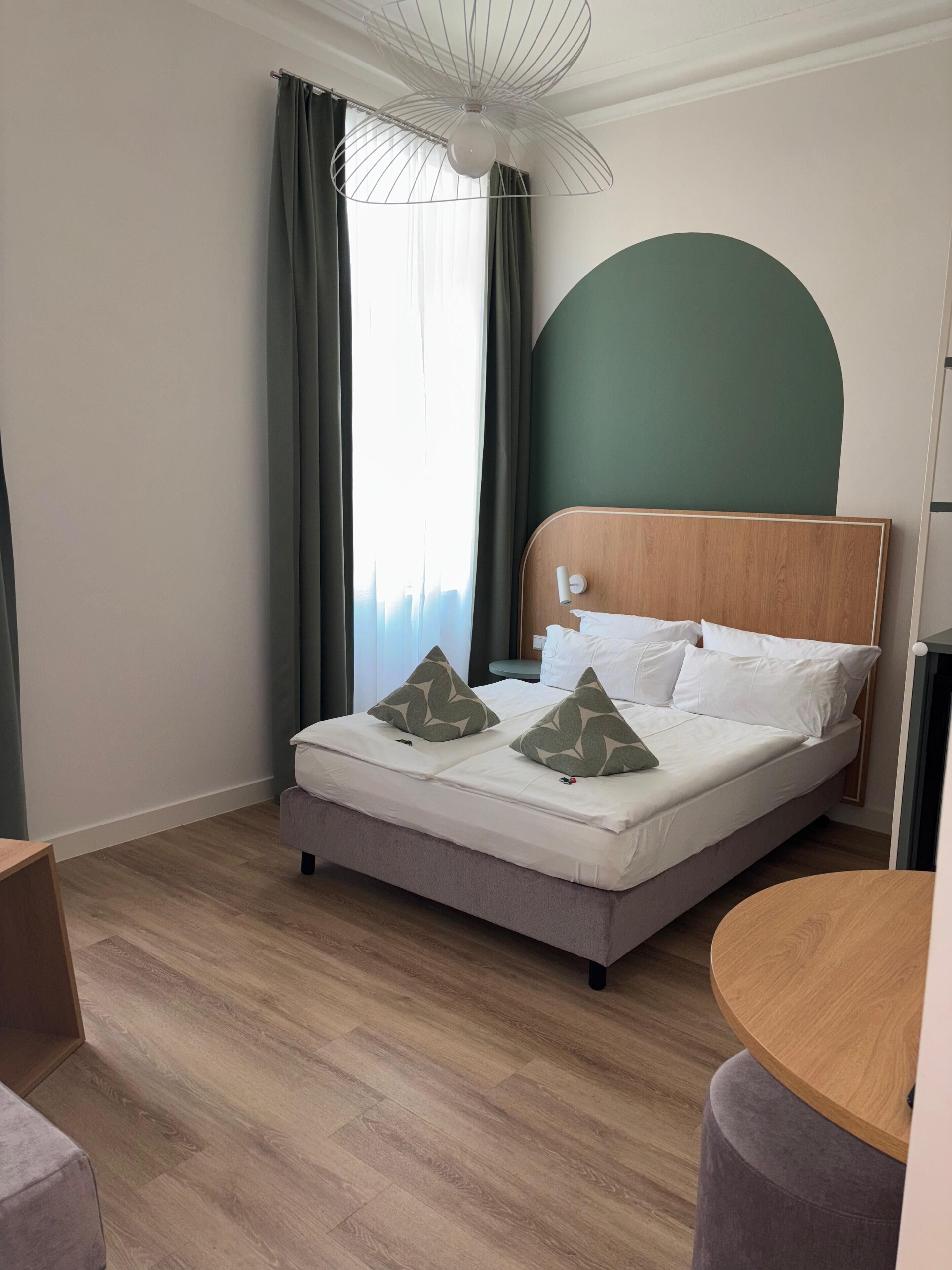 Quarto de hotel moderno com cabeceira de madeira, cama grande com roupa de cama branca e almofadas estampadas, mesa de madeira redonda e vista da cidade através de cortinas leves.