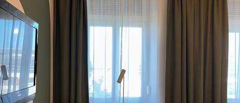 Quarto de hotel com uma cama, uma cadeira, uma escrivaninha e televisão fixada na parede.