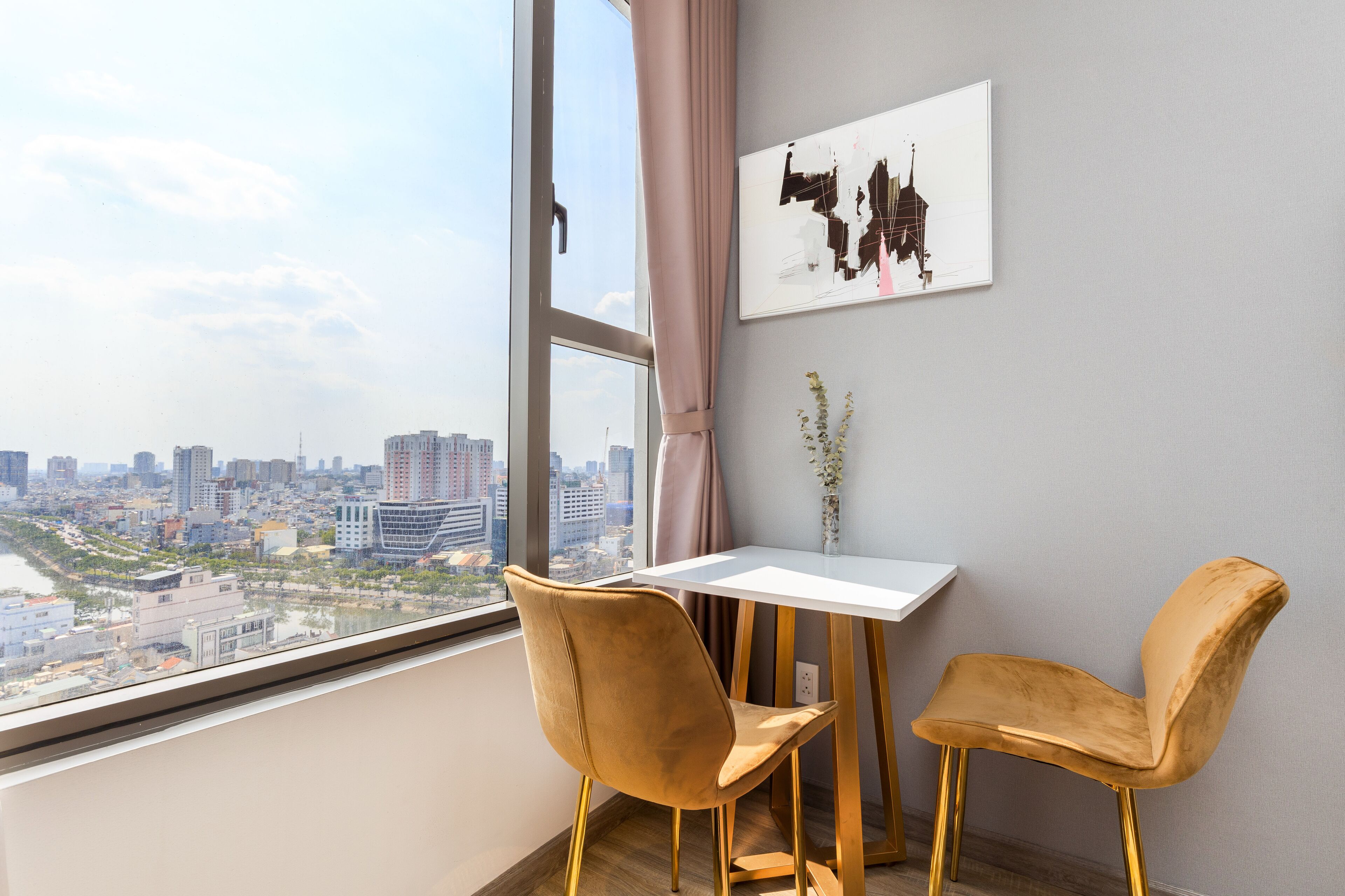 Quarto moderno com vista para a cidade, uma mesa branca, duas poltronas de veludo e uma obra de arte abstrata emoldurada na parede.
