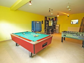 Mesa de bilhar com bolas, mesa de futebol de mesa e área de bar com banquetas e uma televisão.