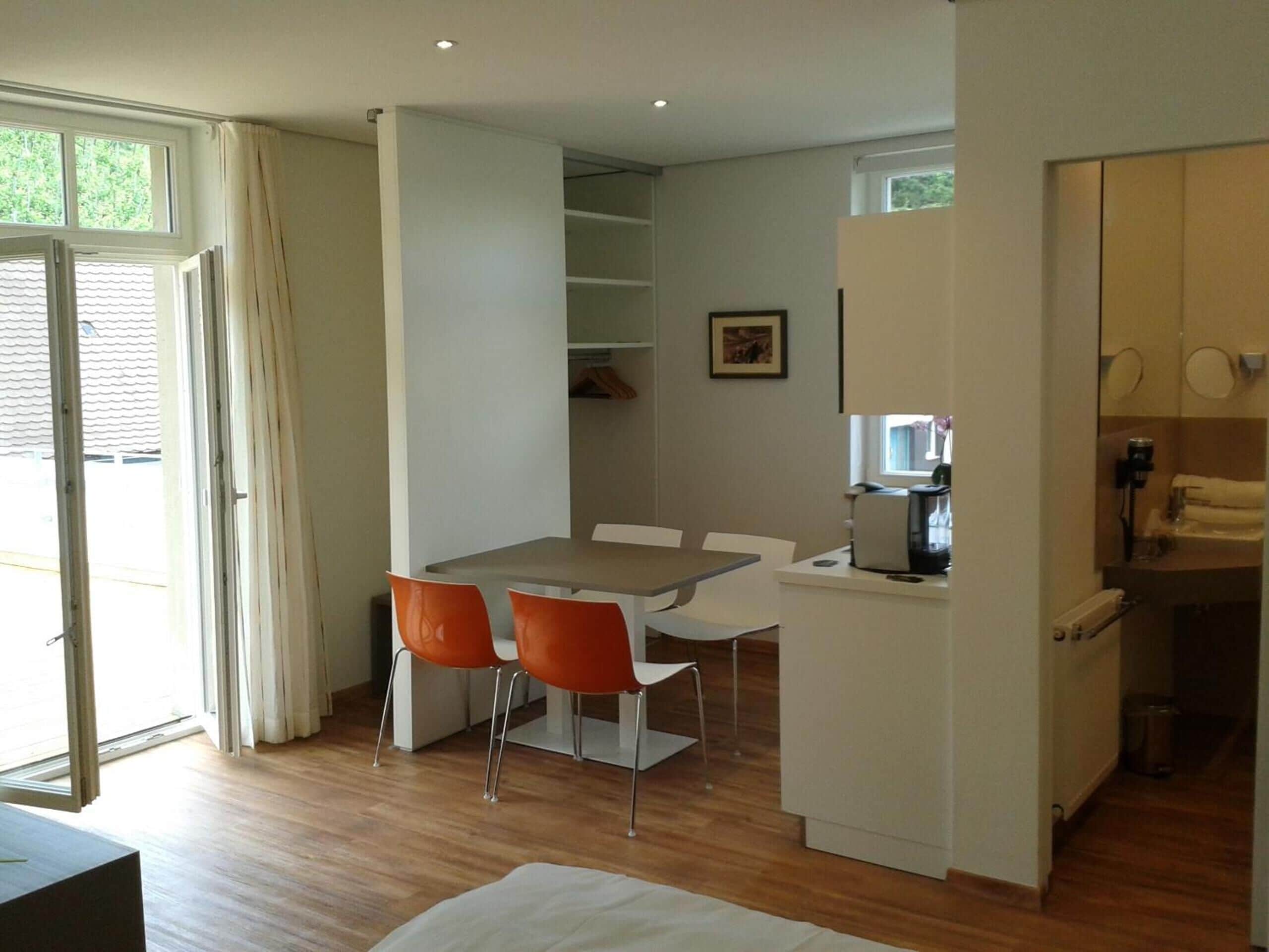Quarto compacto com uma mesa de jantar, duas cadeiras laranja, uma cozinha compacta e uma janela grande.