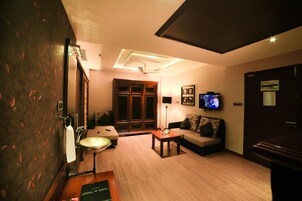 Quarto de hotel com piso de madeira, sofá, mesa pequena, televisão e porta.