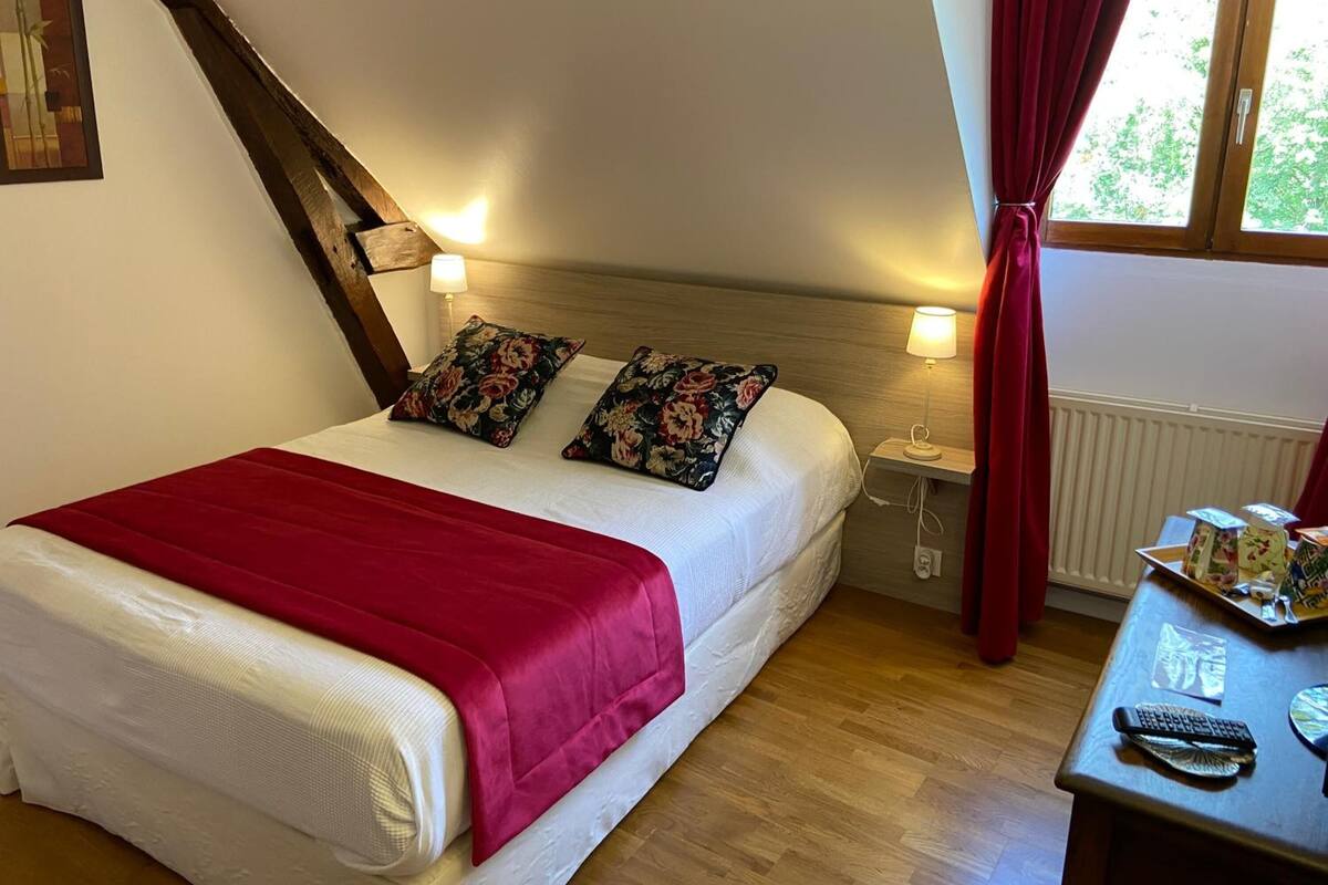 Quarto com teto inclinado, cama com cobertor vermelho, duas almofadas florais, abajures, mesa de cabeceira de madeira e janela com cortinas vermelhas.