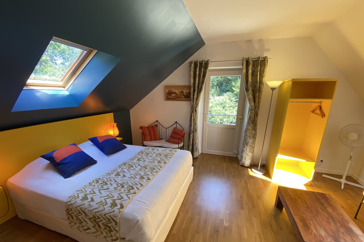 Quarto com cama, mesa de cabeceira, um banquinho, janela e porta.