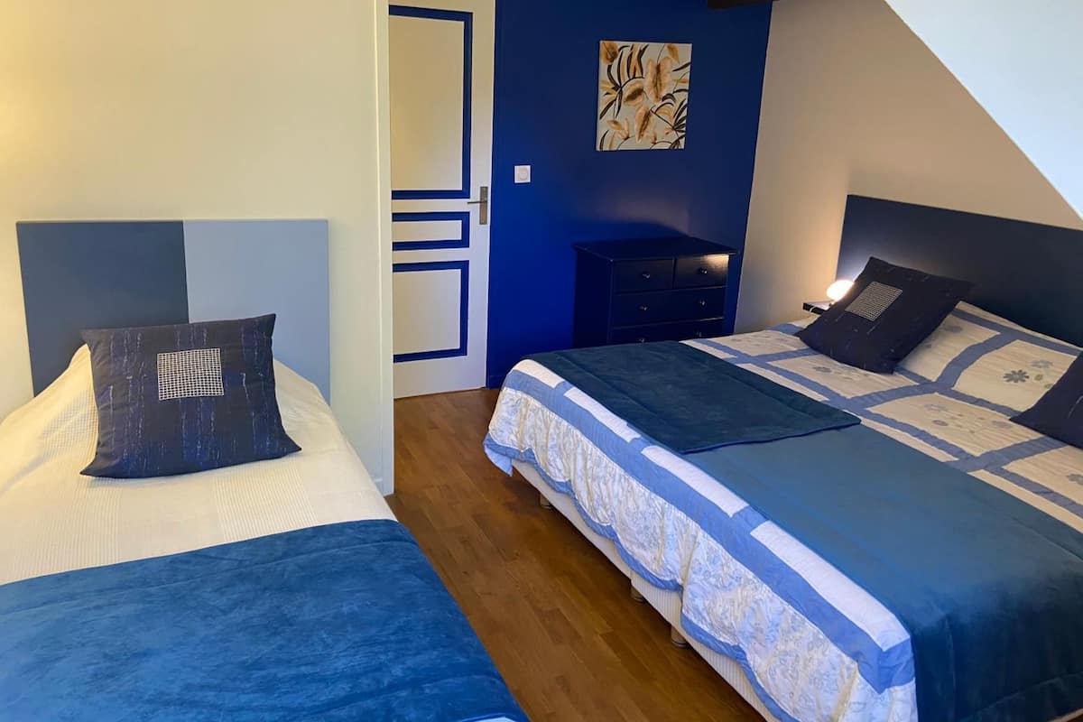 Quarto com duas camas, cada uma com colcha e almofadas azuis. Um criado-mudo com abajur fica entre as camas.