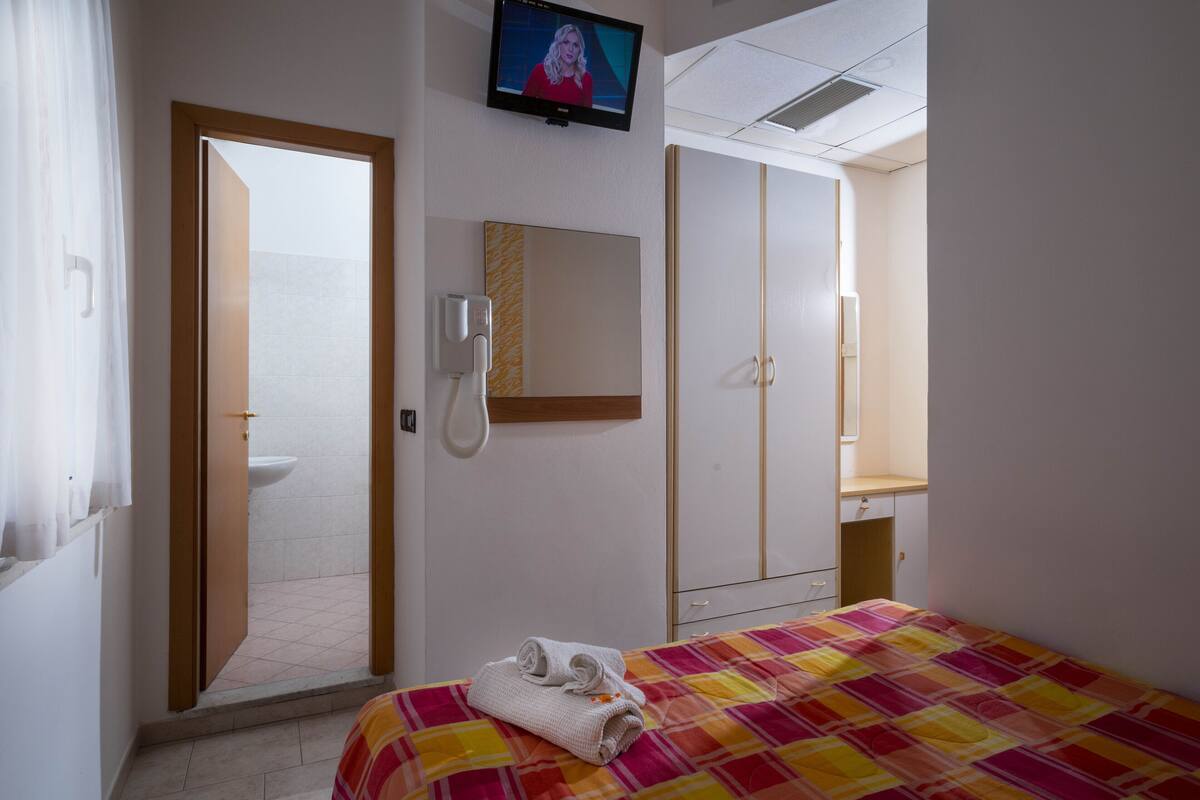 Quarto de hotel com cama, televisão, guarda-roupa e banheiro visível através de uma porta aberta.