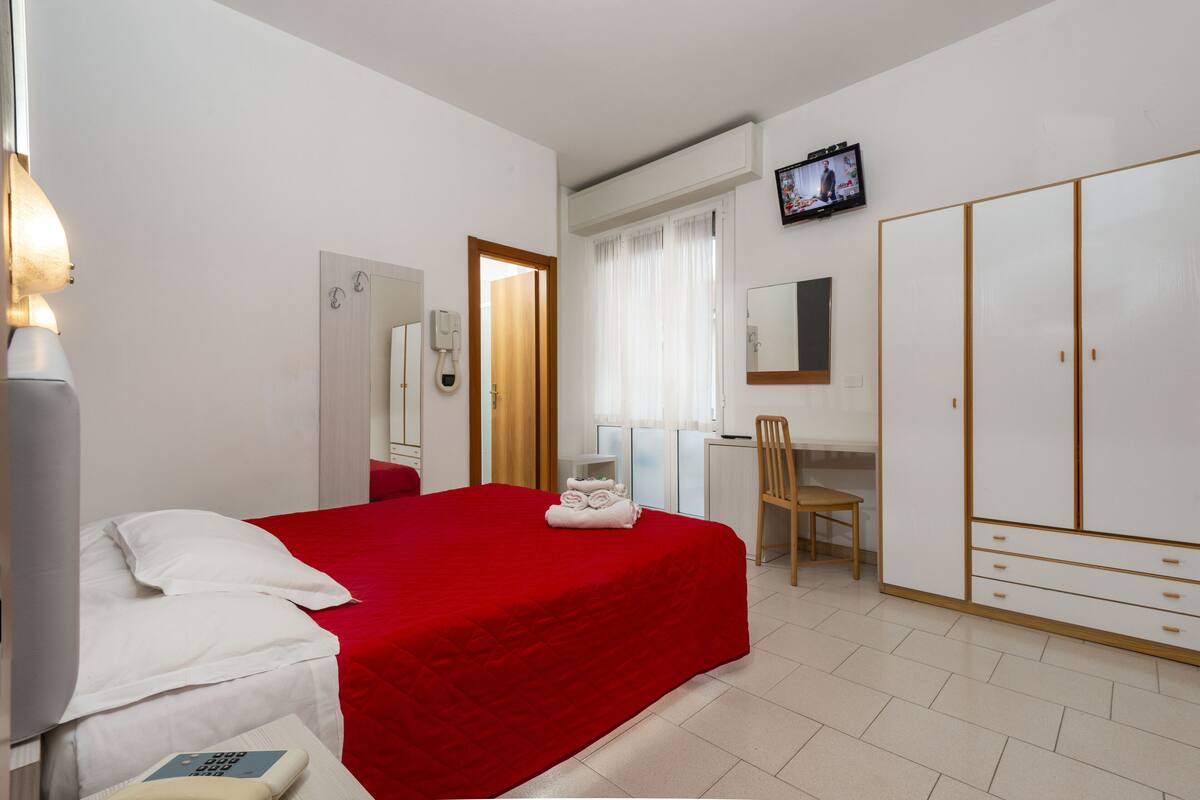 Quarto de hotel com cama vermelha, escrivaninha, televisão e armário.