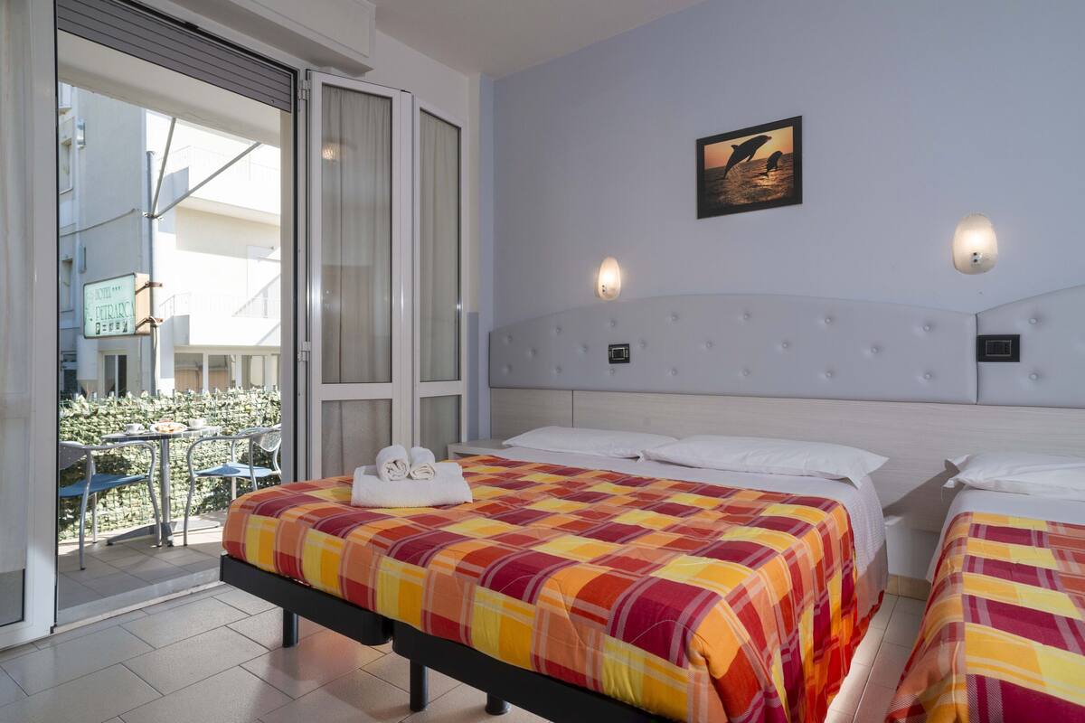 Quarto de hotel com uma cama grande, duas camas menores, uma varanda com mesa e cadeiras, e vista para a cidade.