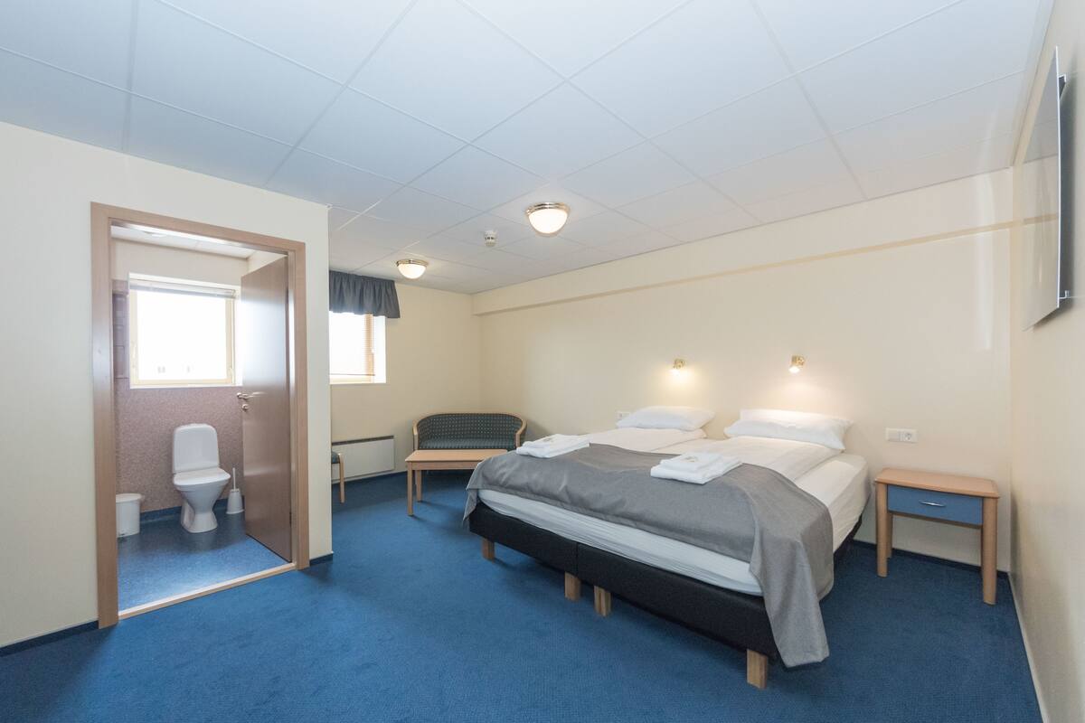 Quarto de hotel com cama, mesas de cabeceira, uma cadeira, sanitário e janela.