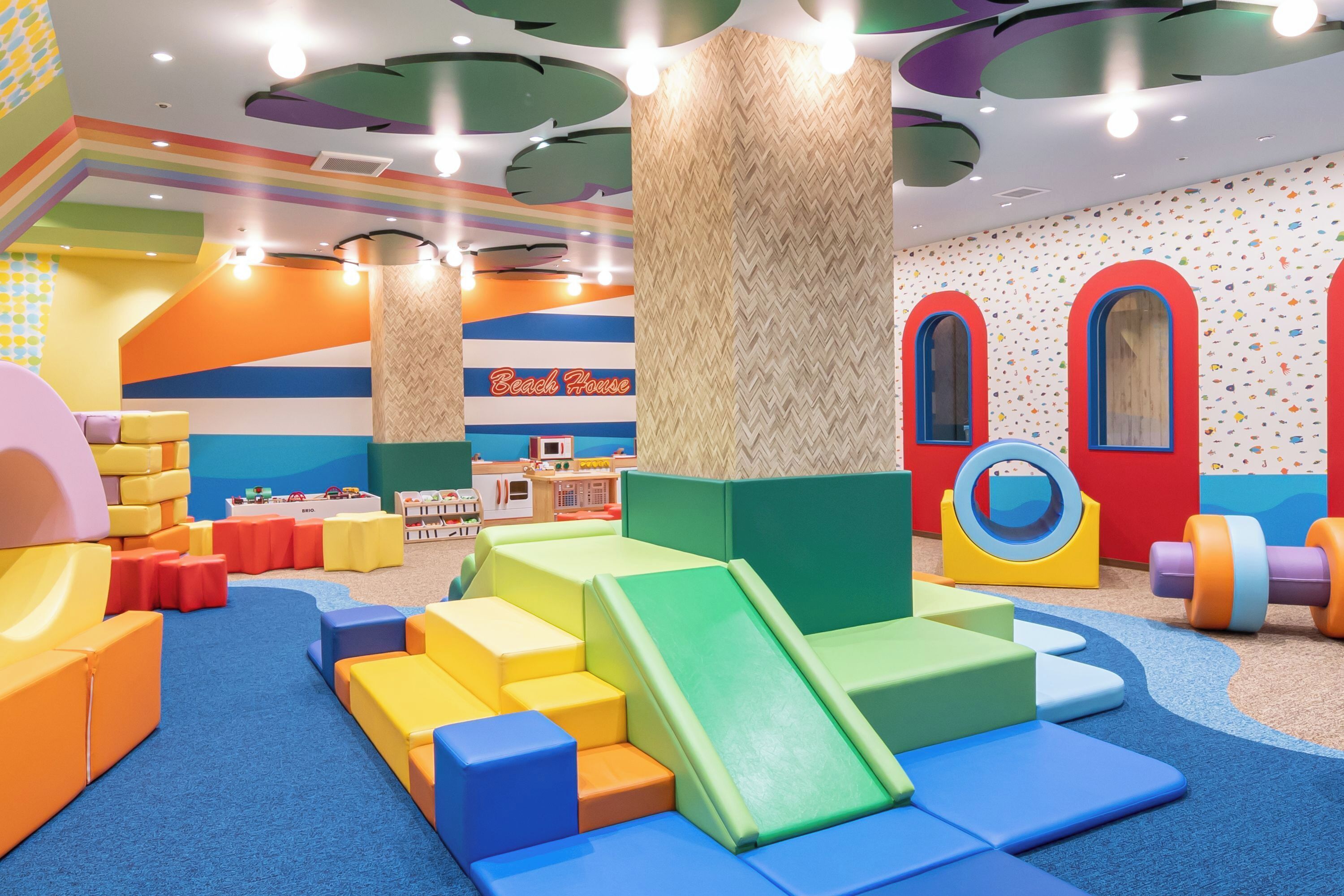 Uma área de recreação interna colorida com estruturas de brincar macias, um cantinho de cozinha e uma parede com o letreiro 'Beach House'.