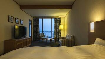 Quarto de hotel com cama, televisão, área de estar com mesa e cadeiras, e vista pela janela.