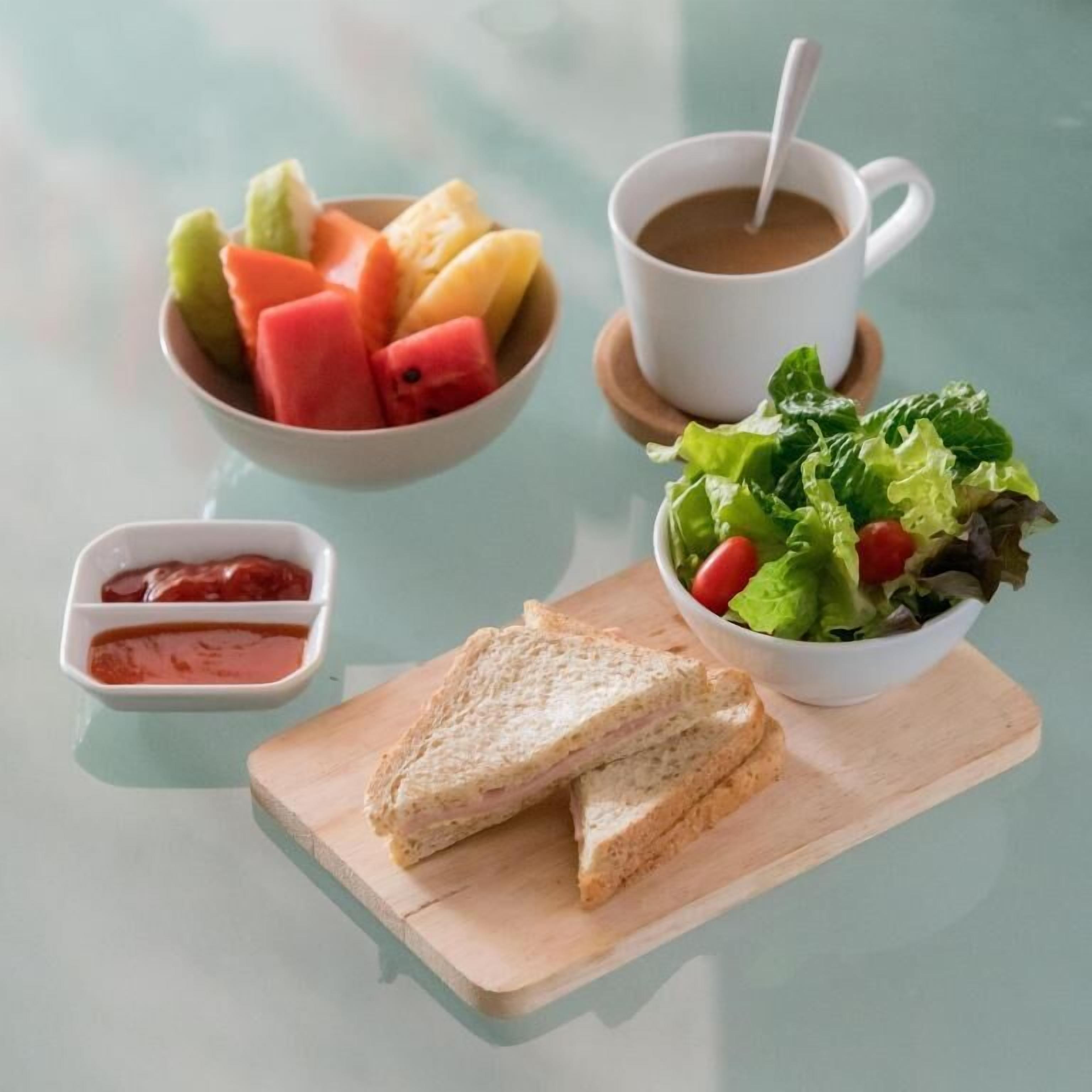 Uma mesa com uma refeição que inclui uma tigela de frutas mistas, uma xícara de café, um pequeno recipiente de ketchup, um sanduíche e uma salada.