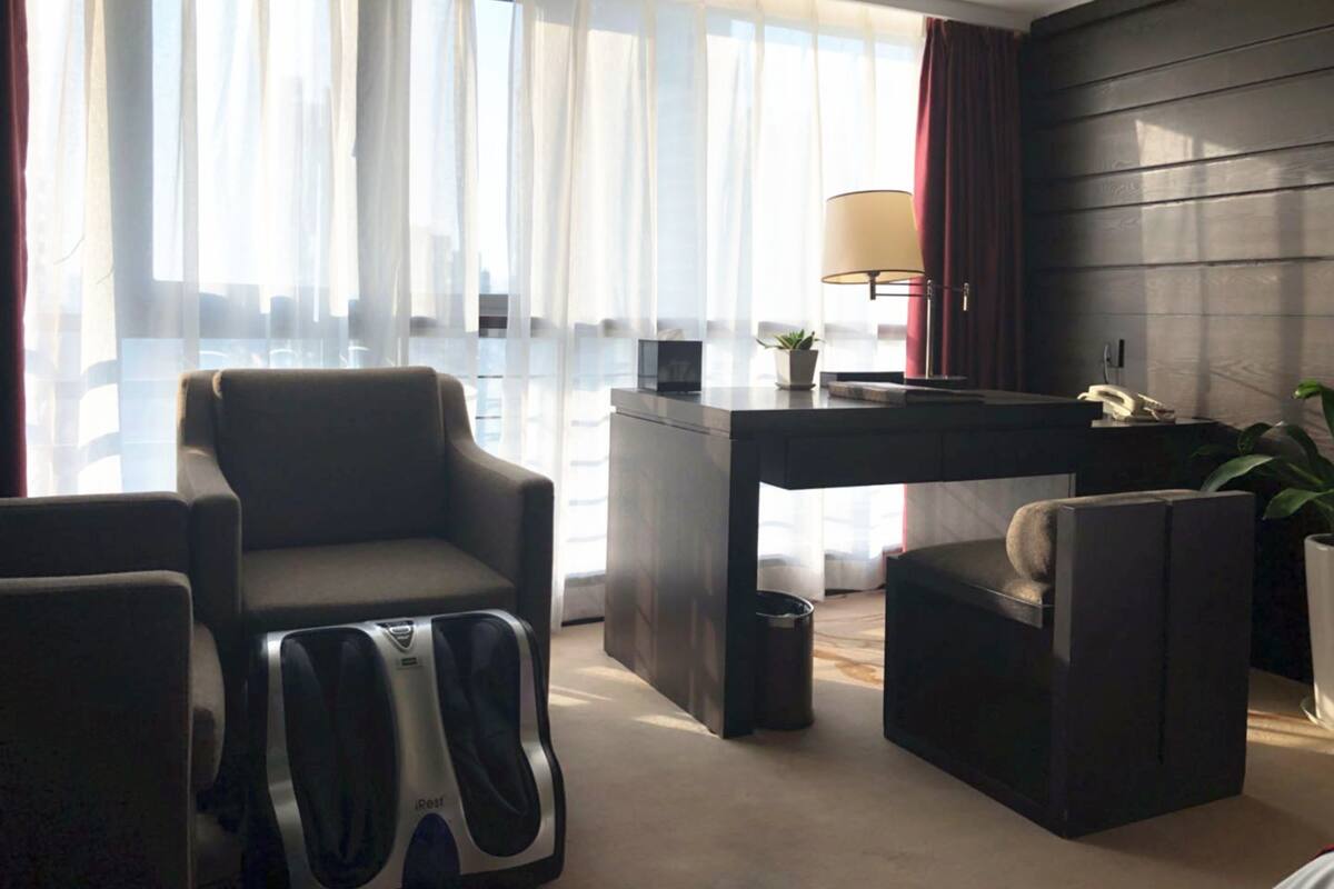 Quarto de hotel moderno com uma janela grande, uma escrivaninha, duas poltronas, uma mesa pequena e um vaso de planta.