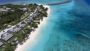 Vista aérea de um resort à beira-mar com bangalôs sobre a água, águas turquesa cristalinas e uma praia de areia.
