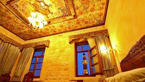 Quarto com cama, um candelabro, vista de uma torre pela janela e teto estampado.