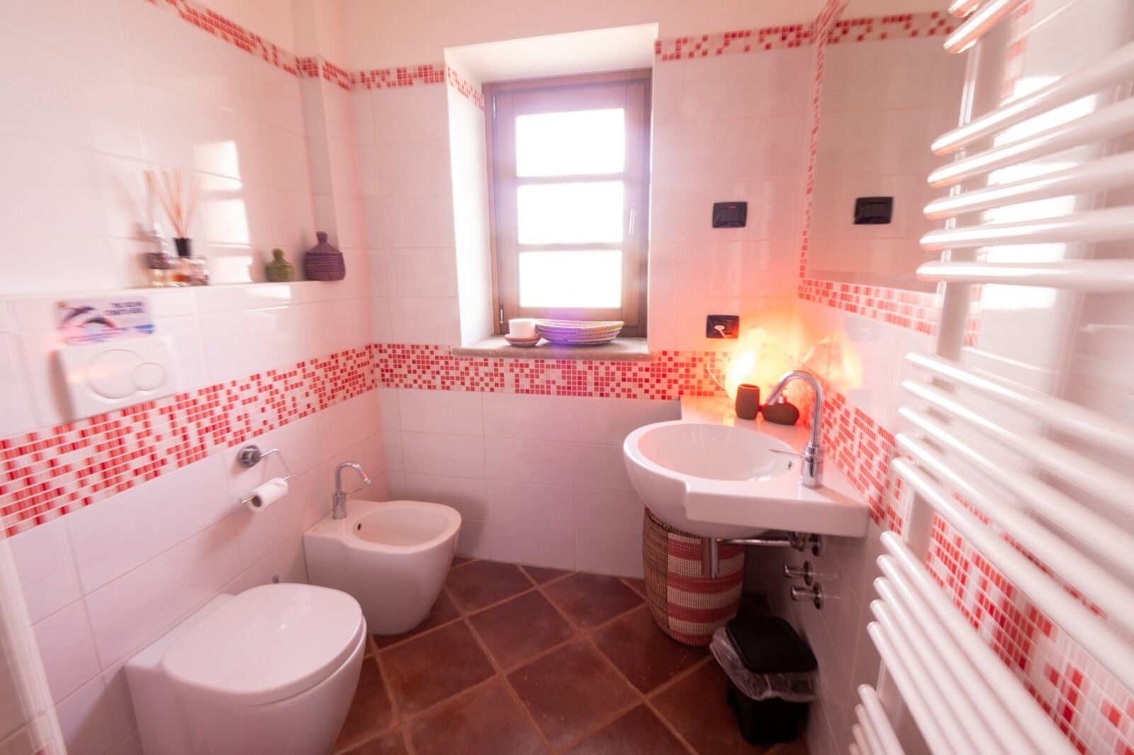 Casa de banho pequena com sanita, bidé e lavatório, revestida com padrões de mosaico vermelho e branco.