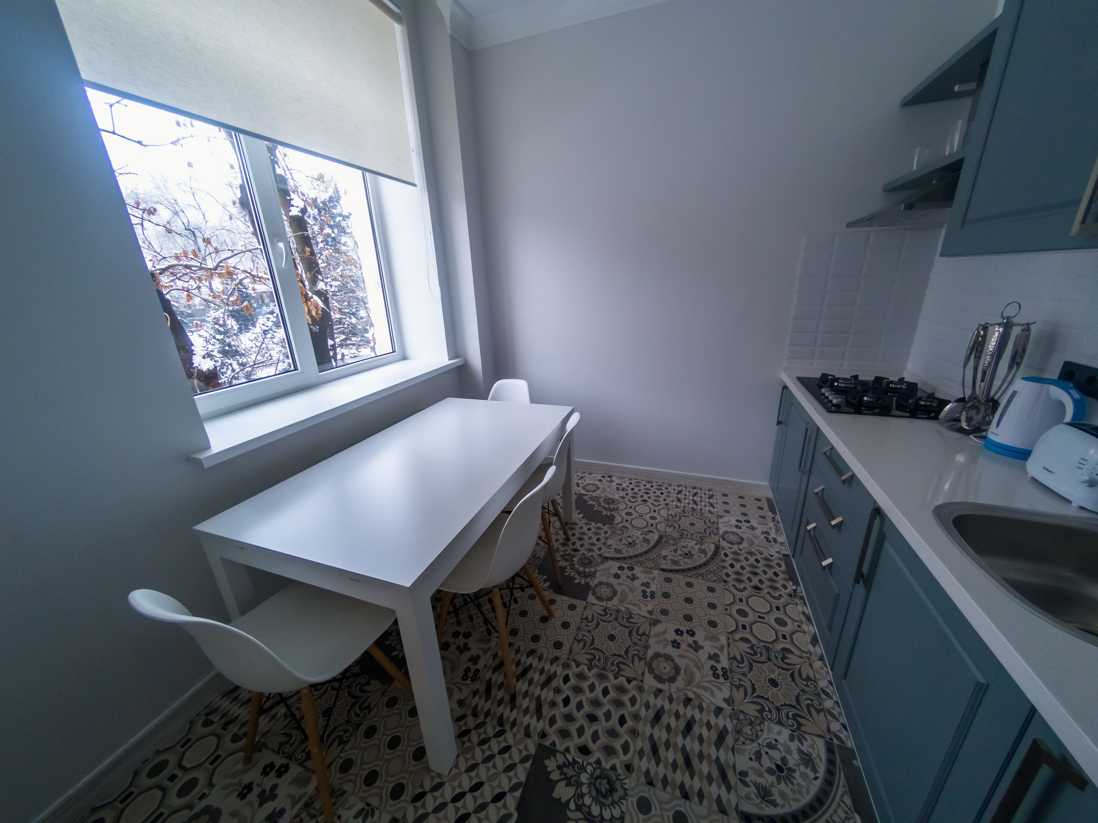 Uma cozinha pequena com mesa e cadeiras brancas, janela com vista para árvores e piso estampado.