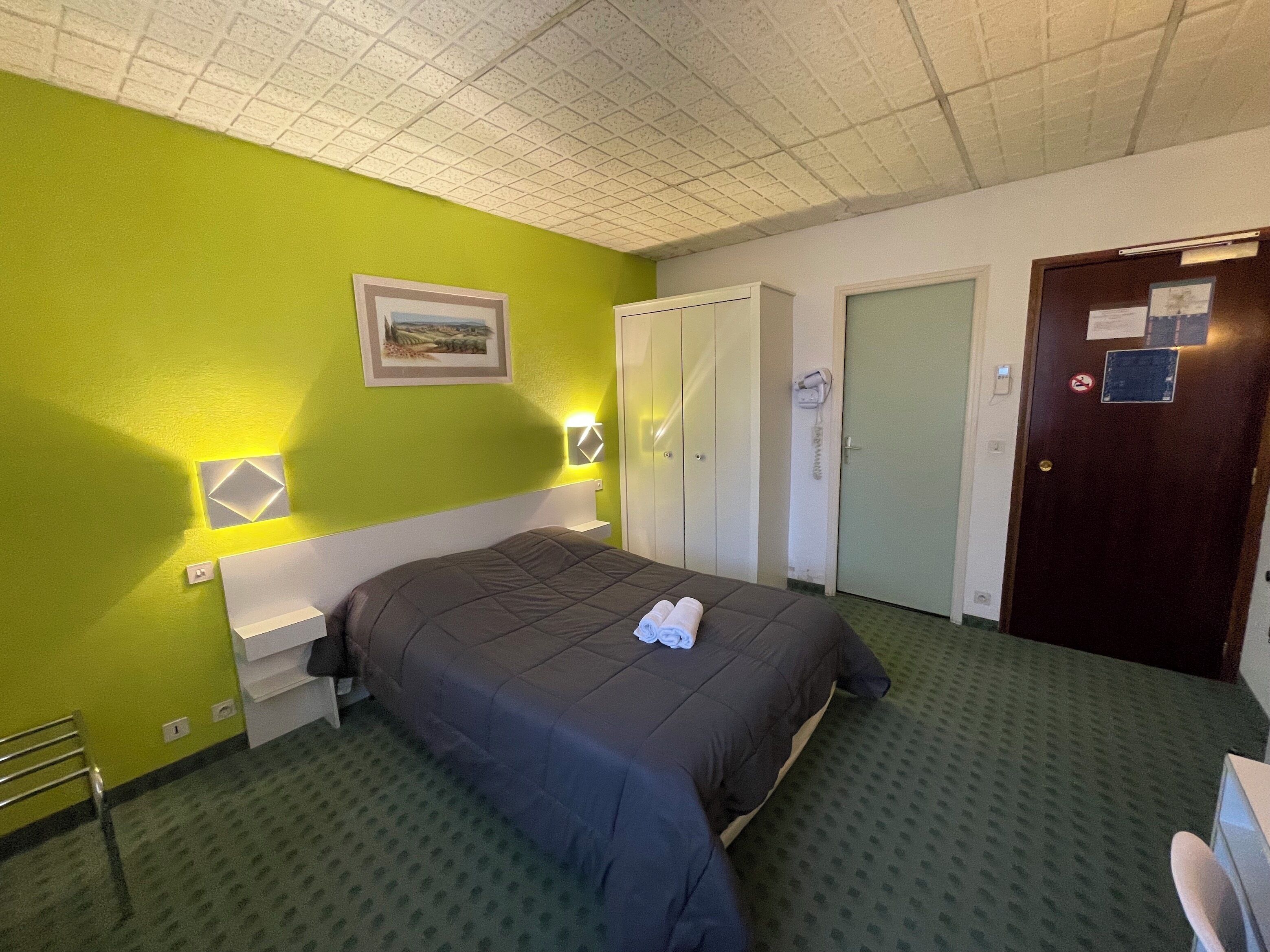 Quarto de hotel com uma cama, parede verde, um quadro e uma porta.